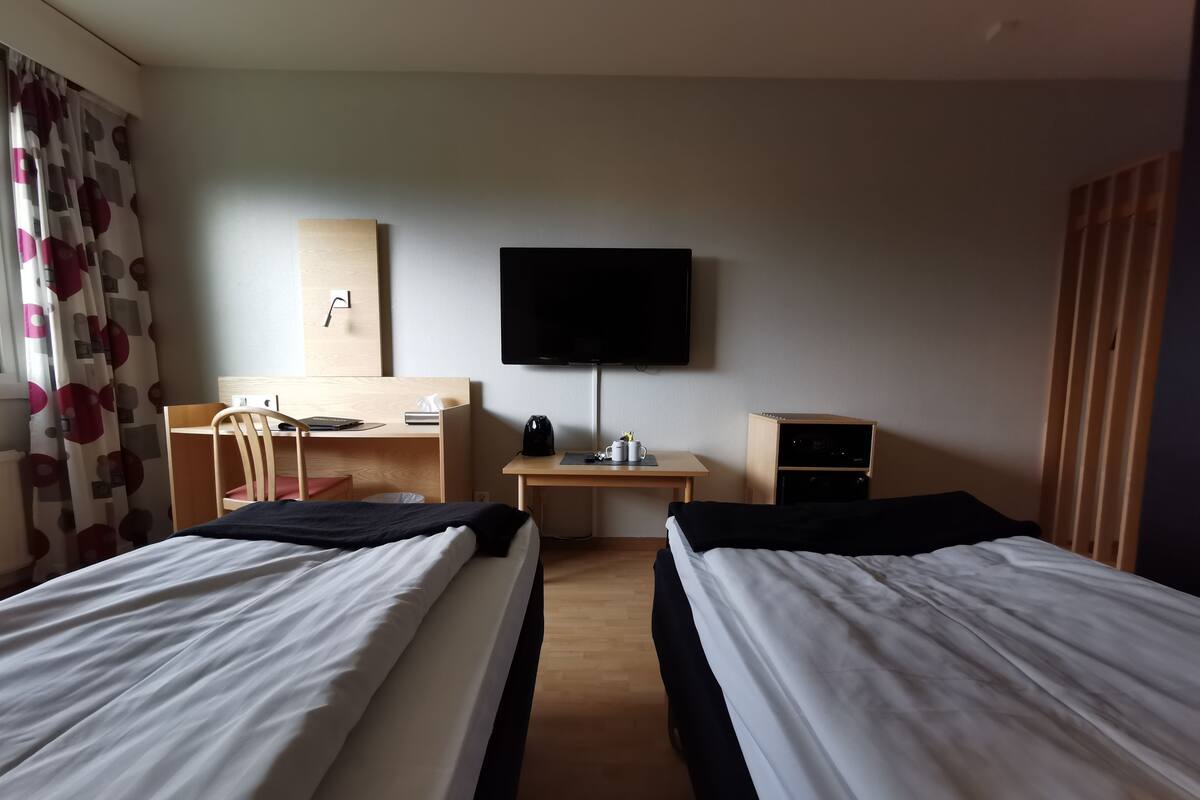 Quarto de hotel com duas camas, uma escrivaninha, uma cadeira, uma televisão e um guarda-roupa.