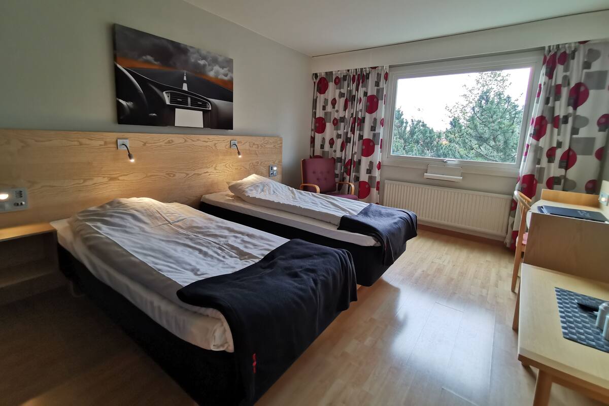 Quarto de hotel com duas camas, uma escrivaninha, uma cadeira, uma janela com cortinas e um quadro na parede.