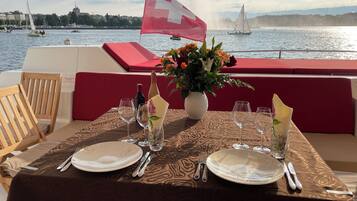 Uma mesa posta para dois, com vista para um lago e veleiros, uma bandeira suíça e um vaso de flores.