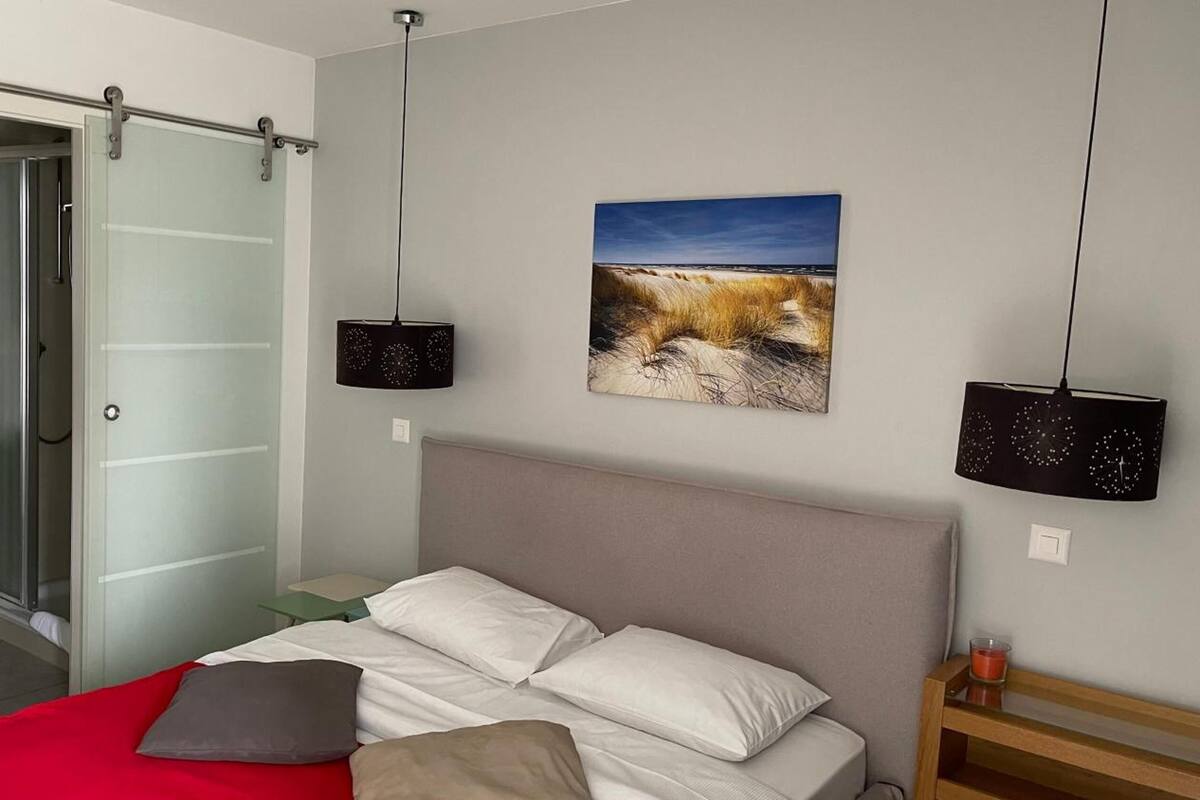 Quarto com cama, manta vermelha, cabeceira cinzenta, pintura de uma praia e duas luminárias pendentes.
