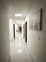 Um corredor comprido e estreito, com paredes brancas, chão de azulejos e portas em ambos os lados.