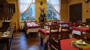 Uma área de refeições com mesas preparadas para uma refeição, uma janela grande com cortinas, e uma estátua de um cavaleiro ao fundo.