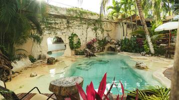 Uma área de piscina com formações rochosas naturais, plantas tropicais e um deck de madeira com cadeiras.