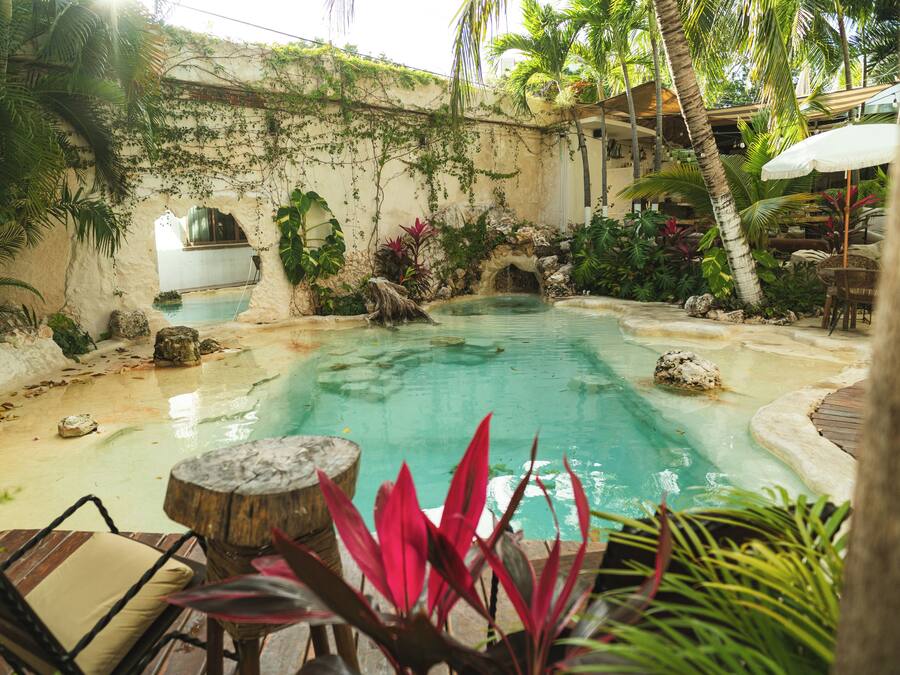 Uma área de piscina com formações rochosas naturais, plantas tropicais e um deck de madeira com cadeiras.