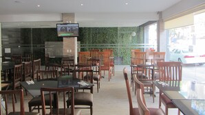 Um restaurante com várias mesas e cadeiras, uma televisão presa a um pilar, e uma parede verde com uma janela que dá para a rua.