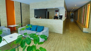 Um hall de hotel moderno com uma mesa de recepção, sofás cinzas, uma mesa de centro e uma grande planta.