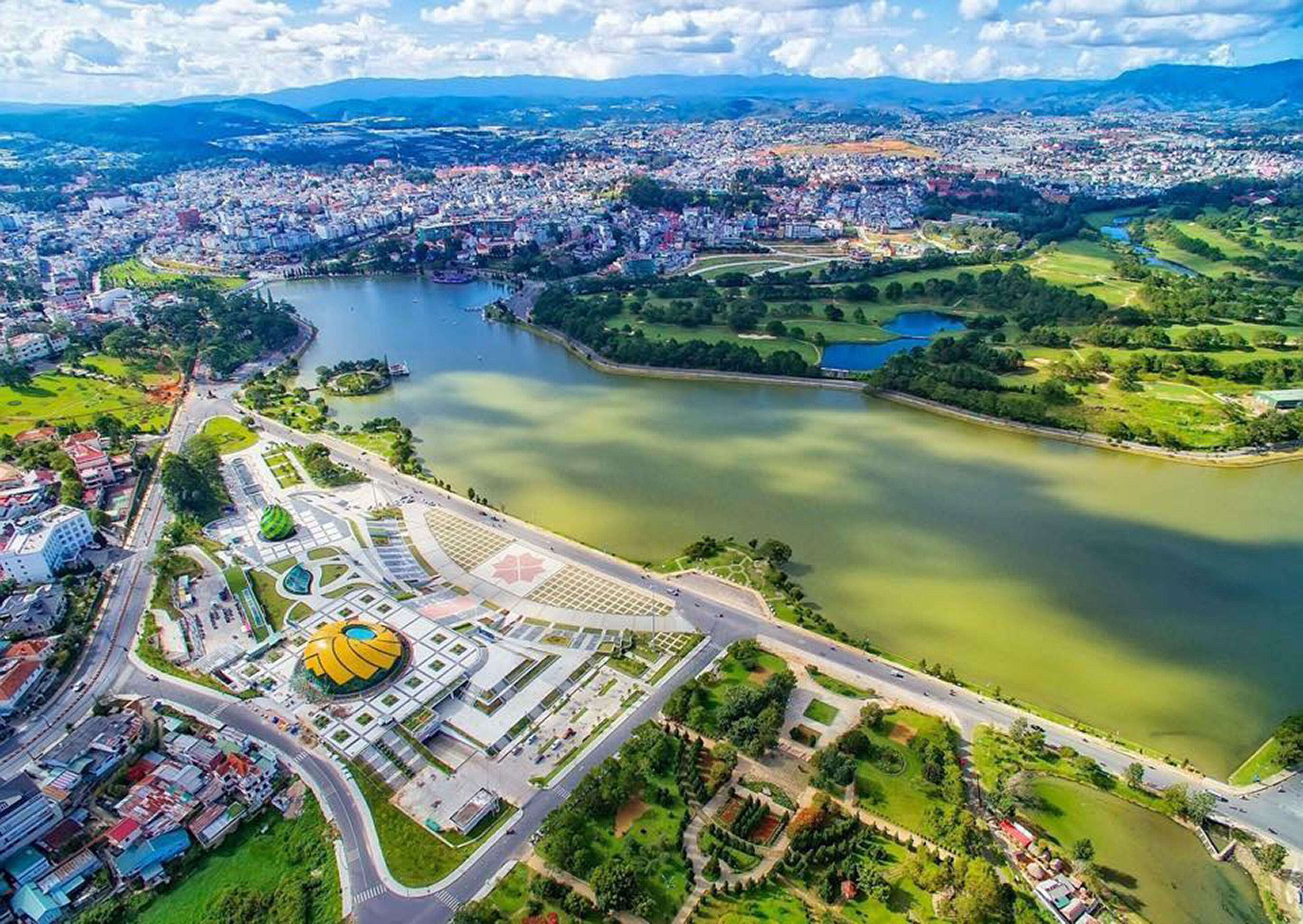 Vista aérea de uma cidade com um grande lago, uma praça central com uma cúpula amarela marcante e áreas verdes ao redor.