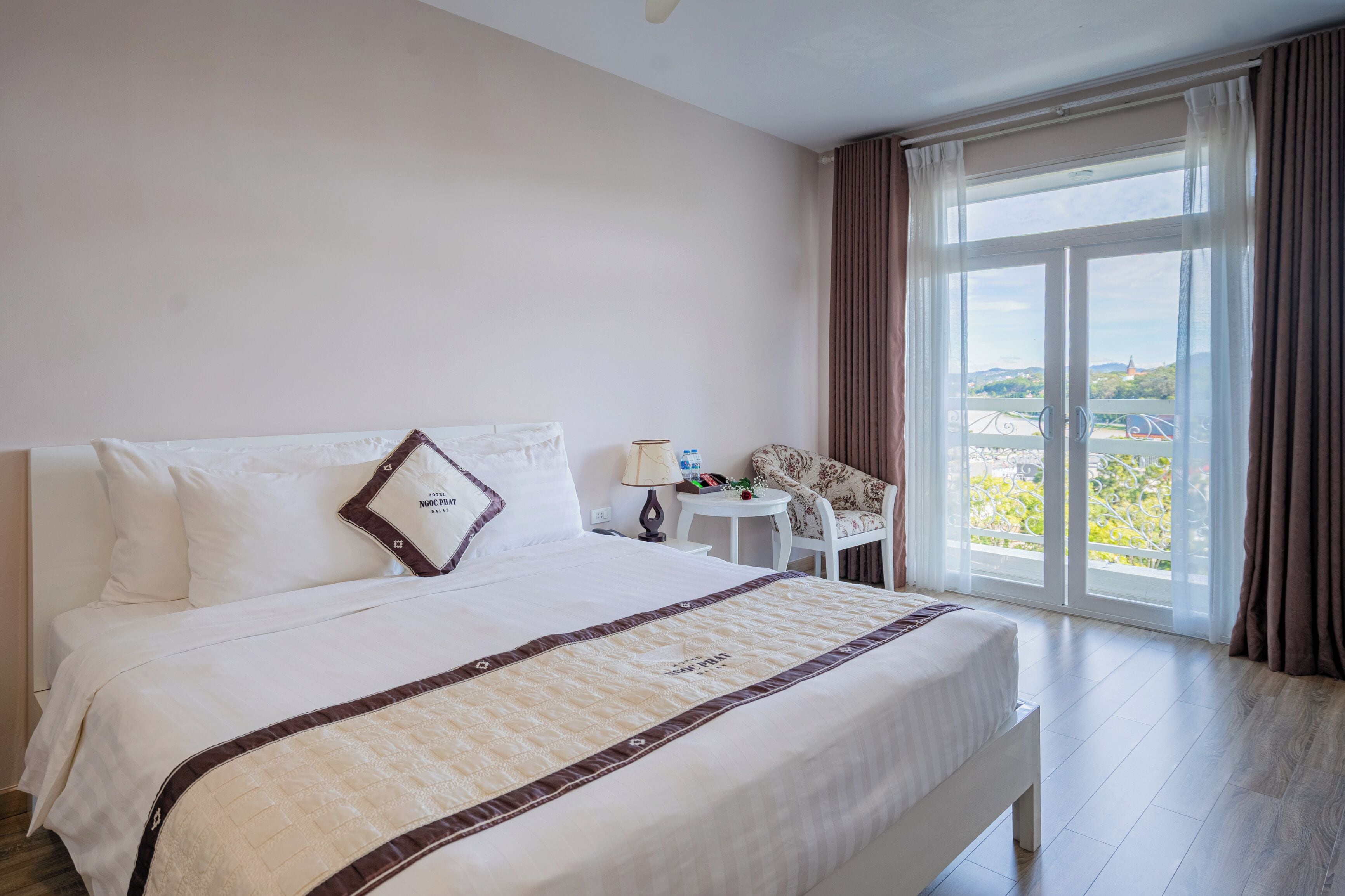 Quarto de hotel com uma cama grande, uma mesinha com abajur e vista para o exterior através de uma janela ampla.