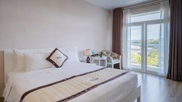 Quarto de hotel com uma cama grande, uma mesinha com abajur e vista para o exterior através de uma janela ampla.