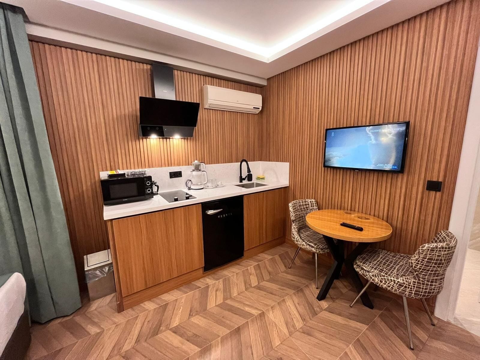 Uma cozinha compacta com micro-ondas, cafeteira e pia, uma pequena mesa de jantar com duas cadeiras e uma TV de tela plana presa na parede.