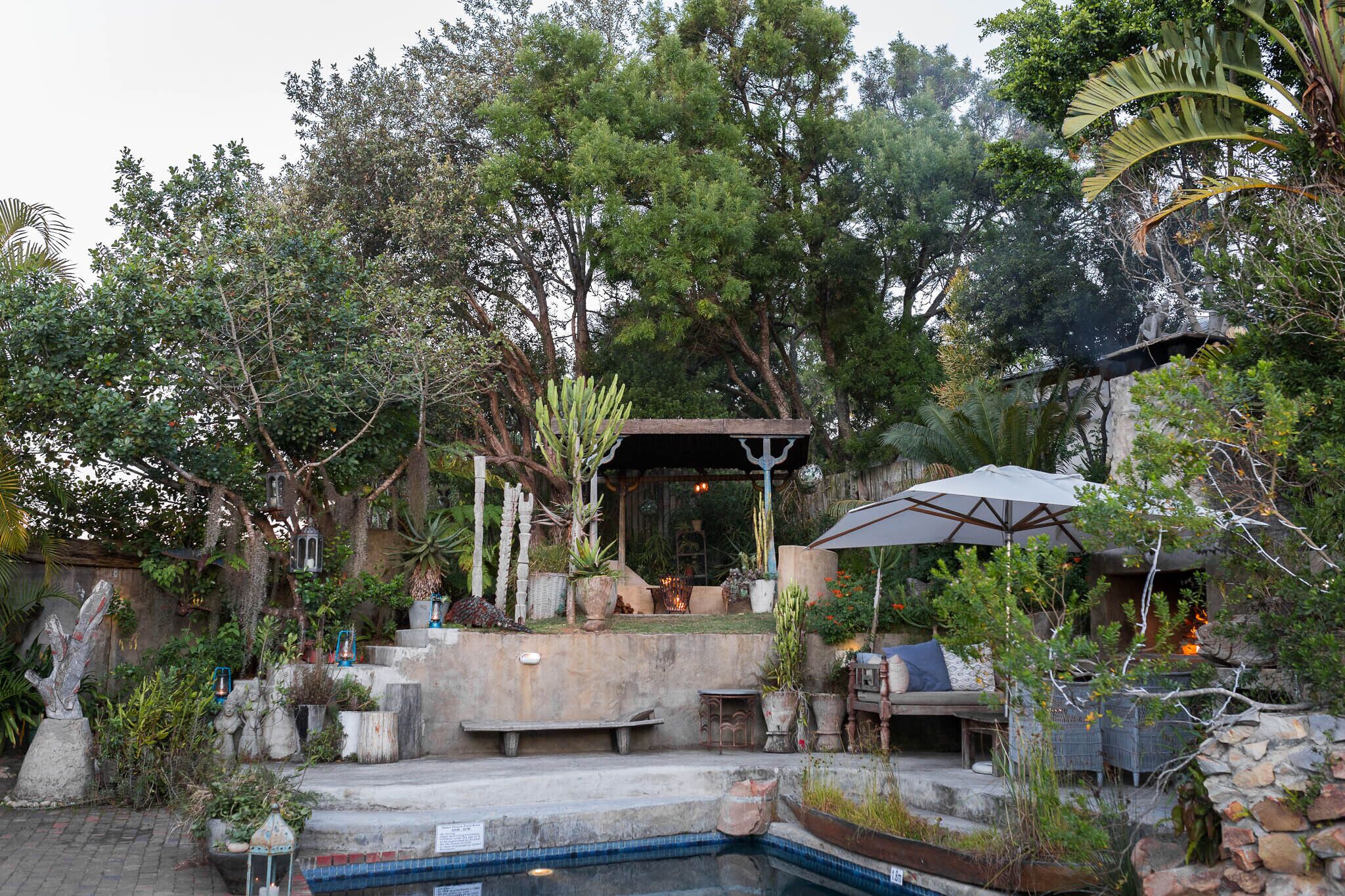 Uma área de piscina com espaço de estar coberto, degraus de pedra e diversas plantas.