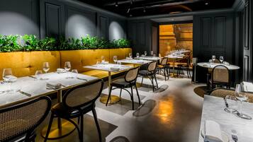 Um restaurante com mesas preparadas para refeições, com uma mistura de cadeiras modernas e clássicas, piso lustroso e parede em tons quentes com vegetação.