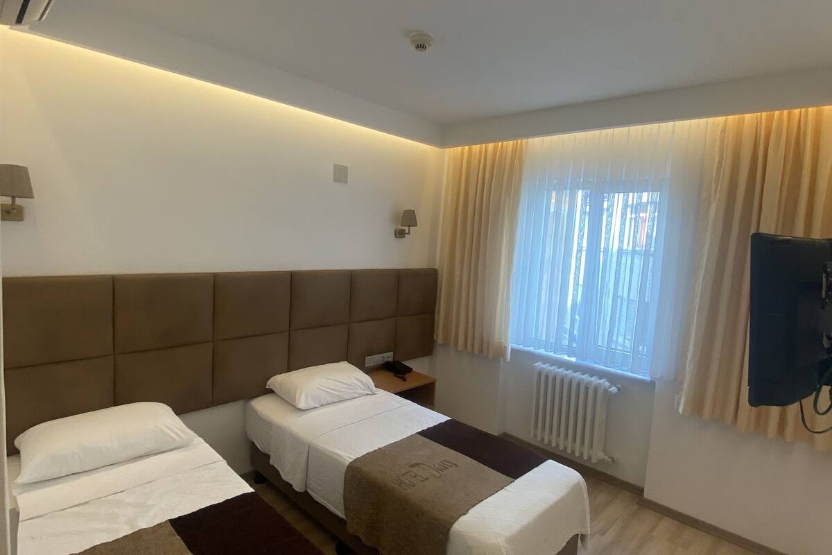 Quarto de hotel com duas camas, uma janela grande com cortinas, uma TV na parede e um radiador.