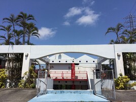 Uma entrada com portão e arco branco, um edifício vermelho e branco ao fundo, e palmeiras.