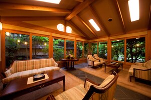 Um quarto espaçoso com teto alto, janelas amplas e vigas de madeira. Possui várias áreas de estar com sofás e cadeiras.