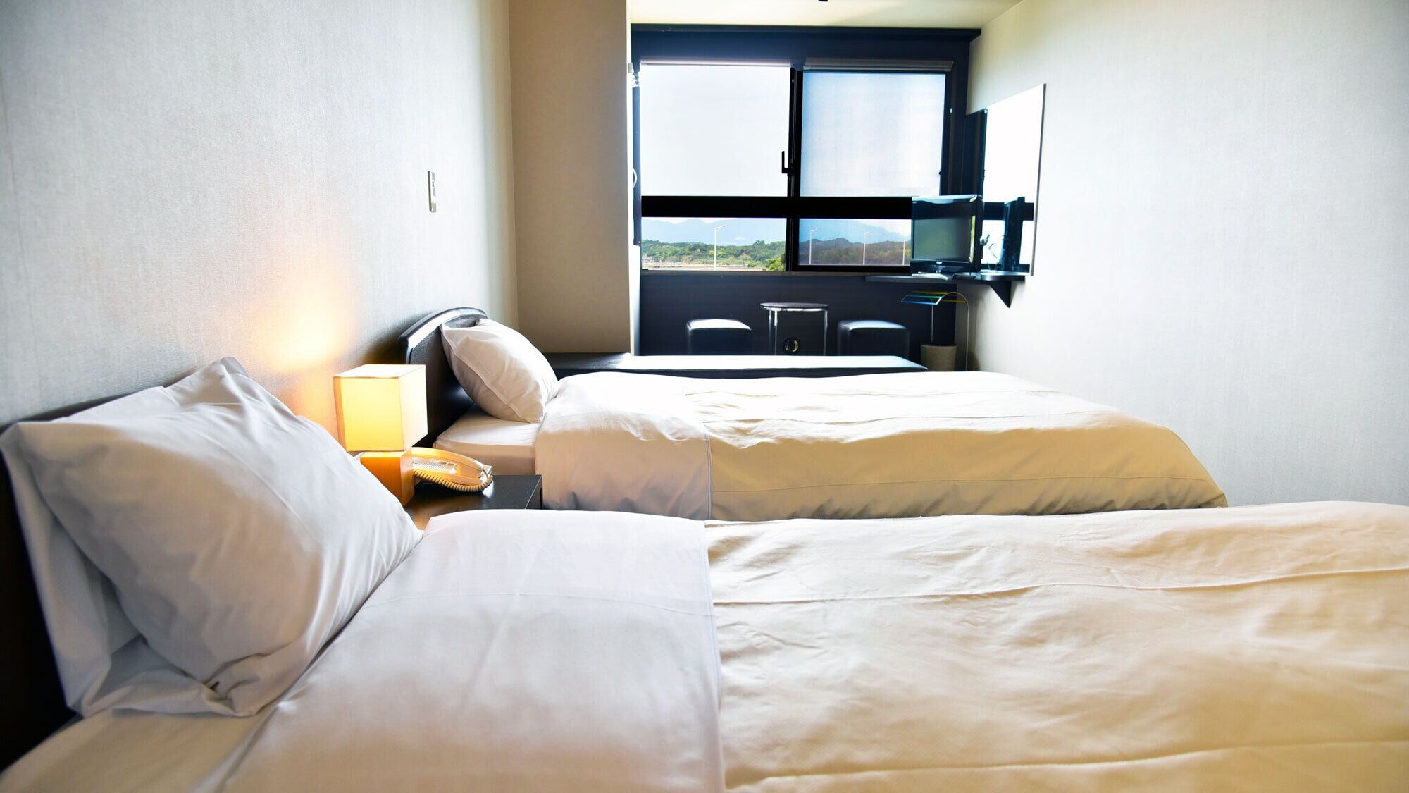 Quarto de hotel com duas camas, uma escrivaninha, uma cadeira, um abajur e uma janela com vista.