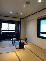 Quarto com tatames, uma mesa baixa com uma cadeira, um sofá e uma janela grande com cortinas.