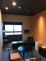 Quarto moderno com escrivaninha, cadeira e uma pequena mesa com almofadas. Há uma janela com persianas, uma TV de tela plana na parede e um radiador no chão.