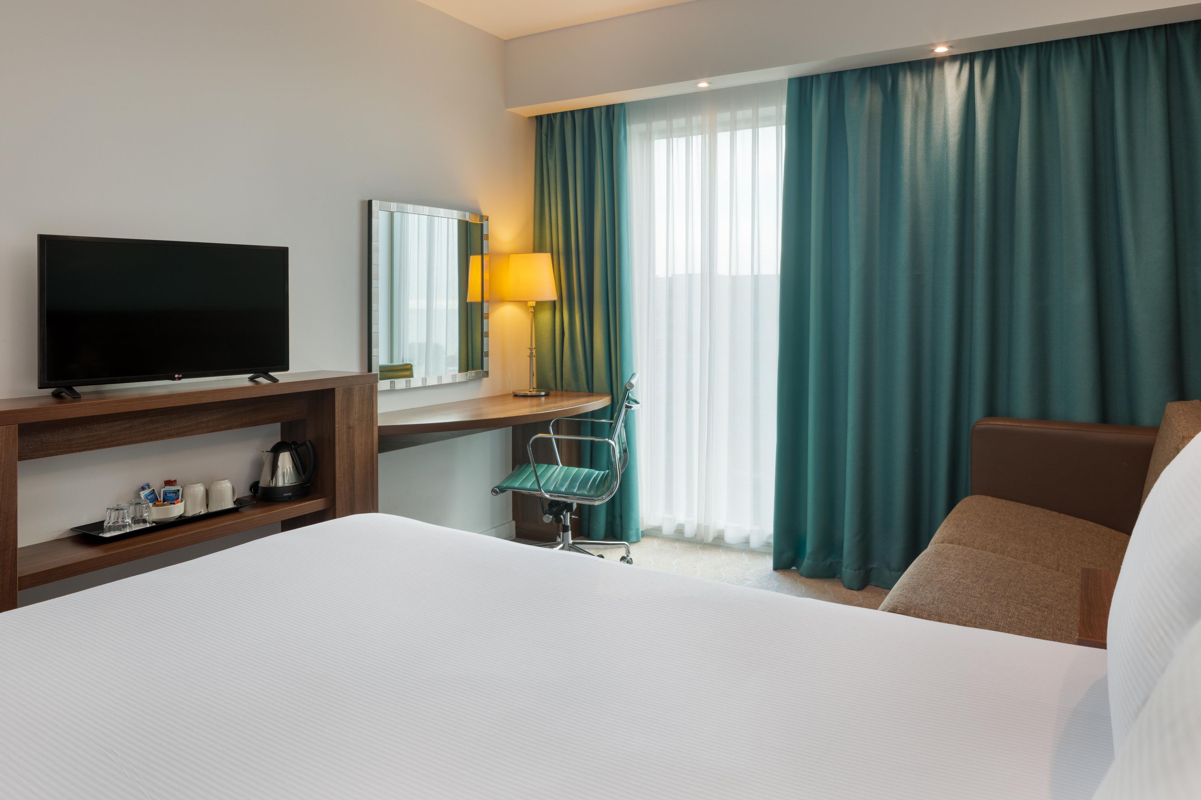 Quarto de hotel com cama, televisão, escrivaninha com abajur, cadeira e janela com cortinas.
