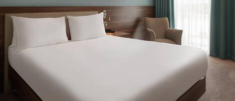 Quarto de hotel com uma cama grande, cabeceira de madeira, mesa de cabeceira, uma cadeira e uma janela com cortinas.