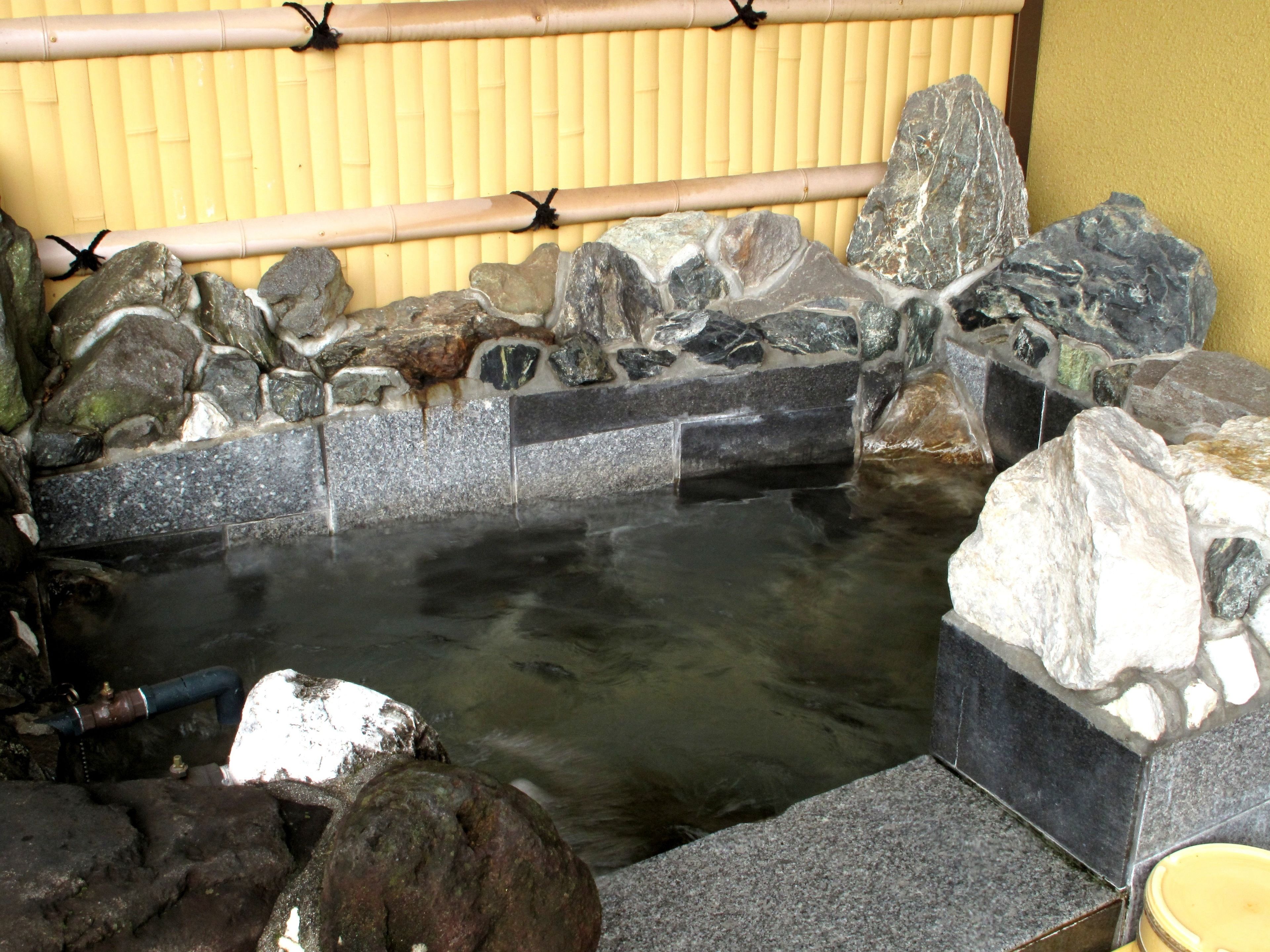 Área de fontes termais com piscina de pedra, rochas e paredes de bambu.