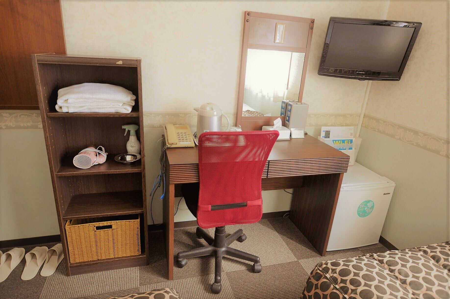 Quarto de hotel compacto com escrivaninha de madeira, cadeira de escritório vermelha, televisão presa à parede, frigorífico pequeno e cama com roupa de cama estampada.