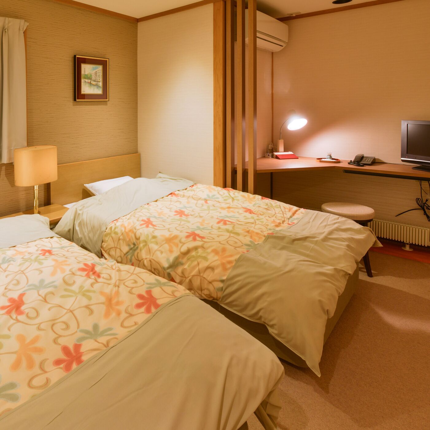 Quarto de hotel com duas camas, uma mesa de trabalho com candeeiro, televisão e aparelho de ar condicionado.