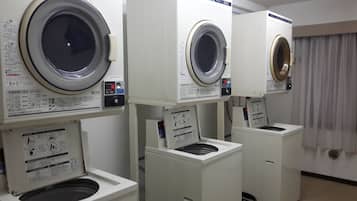 Uma fileira de máquinas de lavar roupa frontais numa lavanderia, com etiquetas de instruções e uma janela com cortinas.