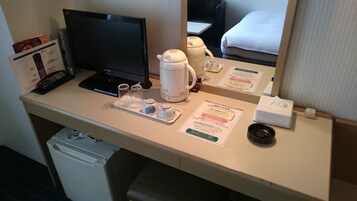 Mesa de quarto de hotel com monitor de computador, cafeteira, bandeja com copos, caixa de lenços e cardápio.
