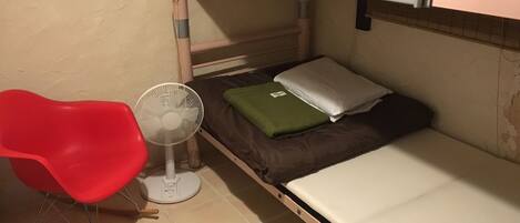 Quarto com beliche, uma cama de solteiro, uma cadeira vermelha e um ventilador.
