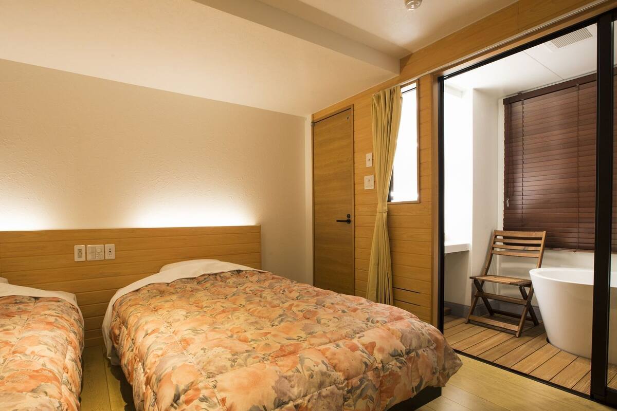Quarto com duas camas, cabeceira de madeira, janela com cortinas, uma cadeira de madeira e uma banheira.