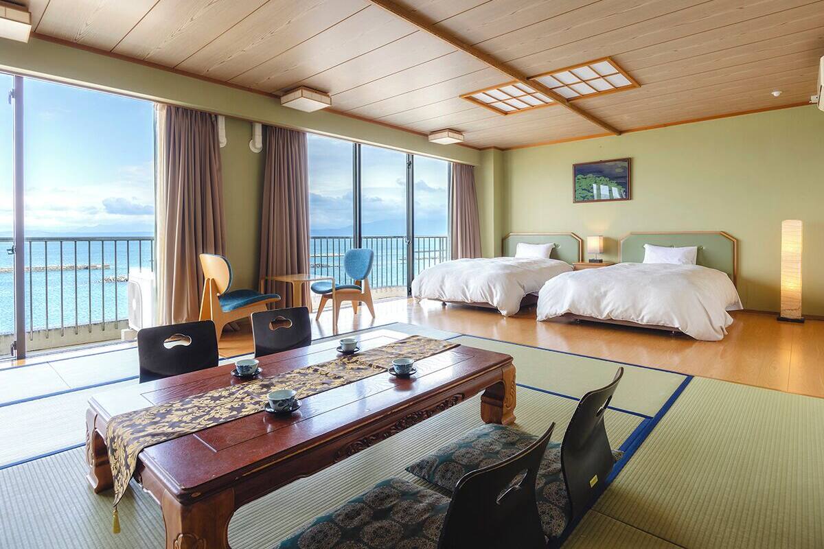 Quarto de hotel com uma janela grande, duas camas, uma mesa de jantar e tapetes tradicionais japoneses de tatame.