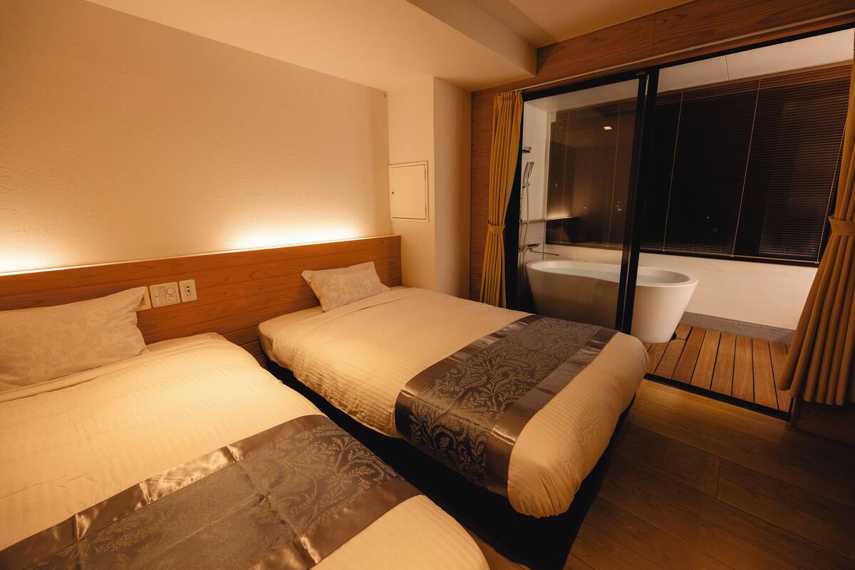 Quarto de hotel com duas camas, cabeceira de madeira, uma janela grande com cortinas e vista para o exterior.