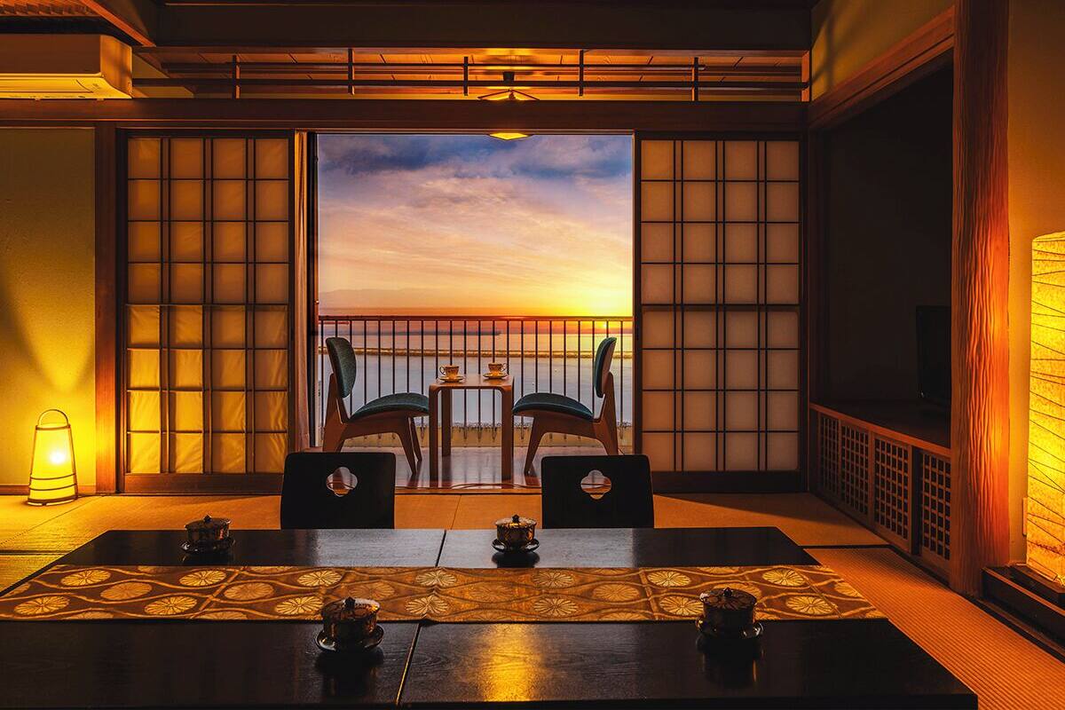 Um quarto japonês tradicional com tatames, uma mesa baixa com um conjunto de chá e vista para o pôr do sol sobre a água.