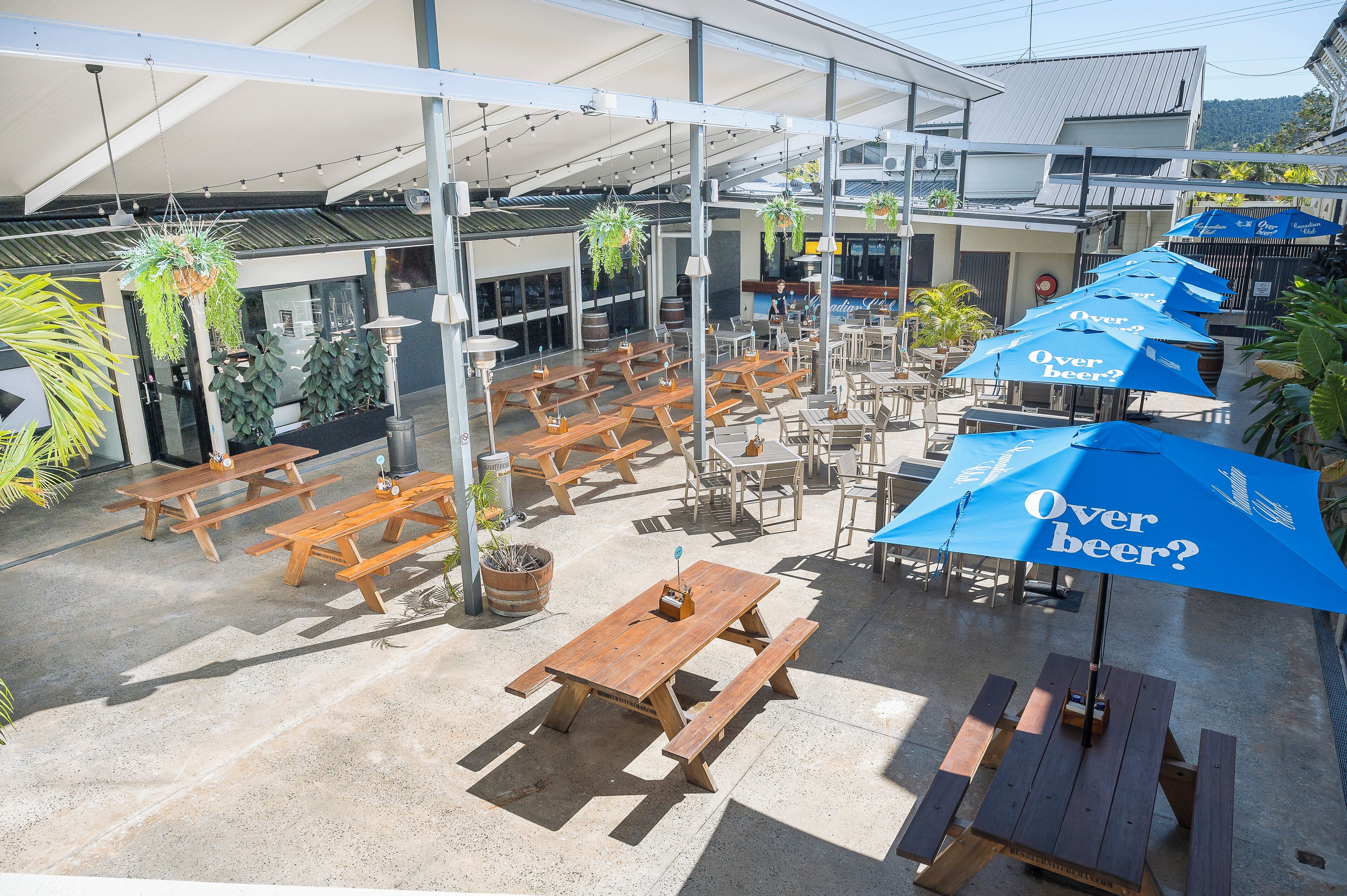 Espaço de pátio ao ar livre com mesas de piquenique de madeira e guarda-sóis azuis com o texto 'Over beer?'.