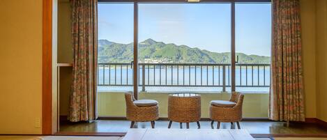 Quarto com uma grande mesa de madeira, duas cadeiras de vime e vista para um lago e montanhas através de amplas janelas.