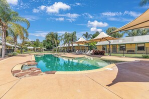 Uma área de piscina com espreguiçadeiras e guarda-sóis, rodeada por edifícios com fachada amarela e detalhes verdes.
