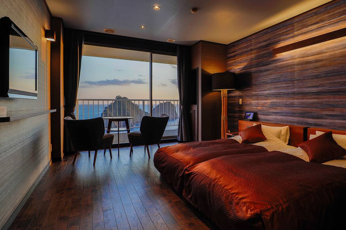 Quarto de hotel moderno com uma cama grande, varanda com mesa e cadeiras, e vista para o mar.