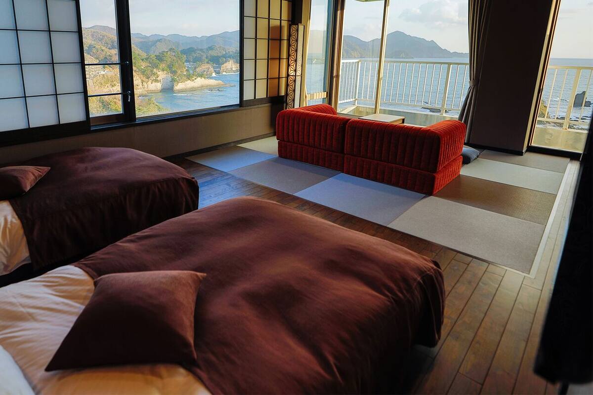 Quarto com duas camas, um sofá vermelho, esteiras de tatame e uma janela grande com vista para o mar e montanhas.