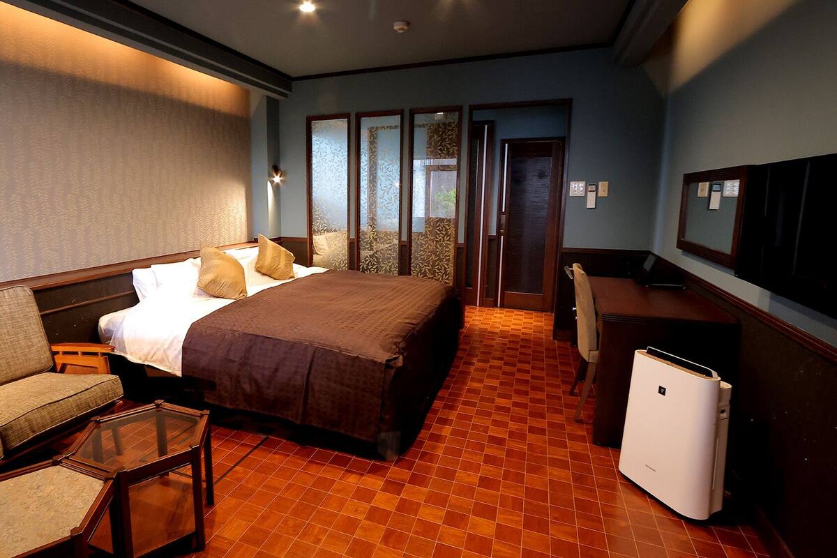 Quarto de hotel com cama, sofá, pequena mesa, escrivaninha e geladeira.