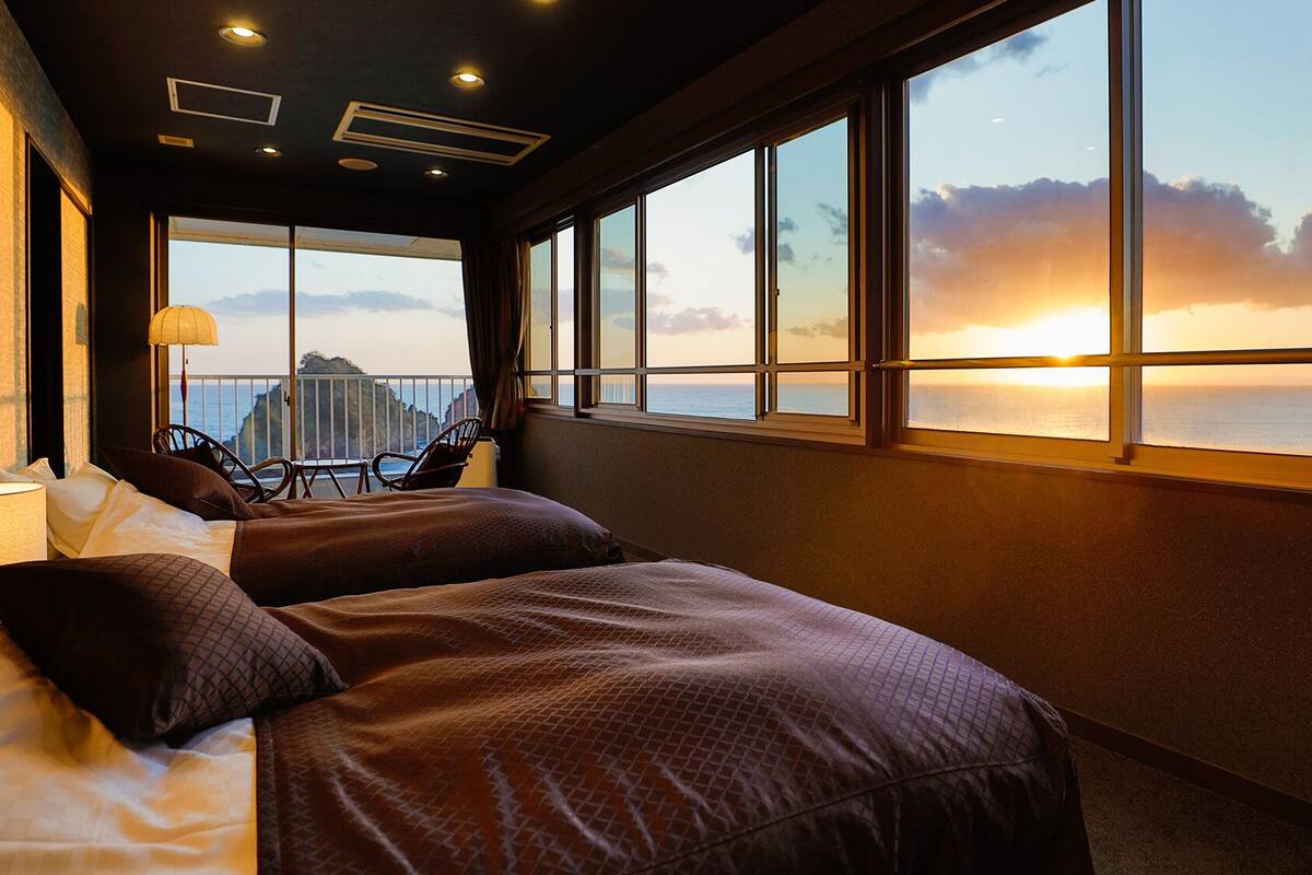 Quarto com cama, cadeira, luminária e uma grande janela com vista para o pôr do sol.