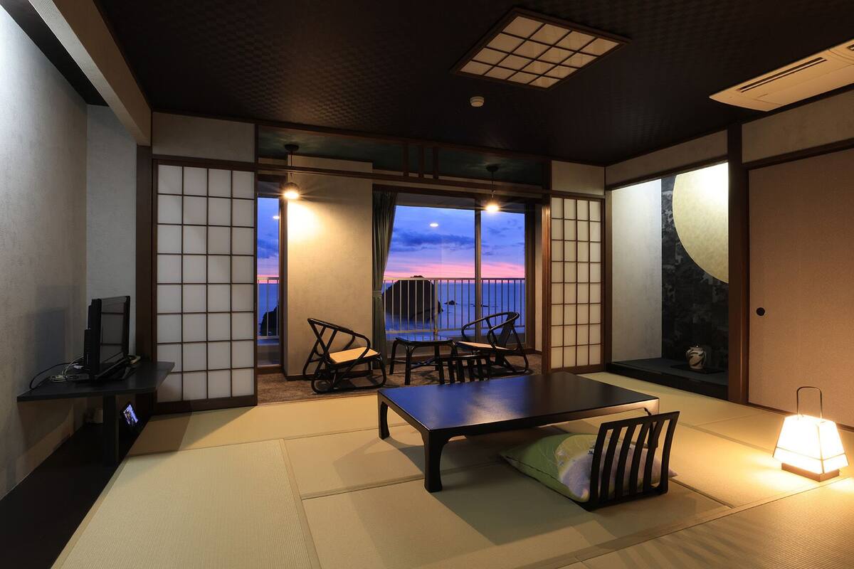 Um quarto japonês tradicional com tatames, uma mesa baixa, cadeiras, uma televisão e vista para o pôr do sol por uma grande janela.
