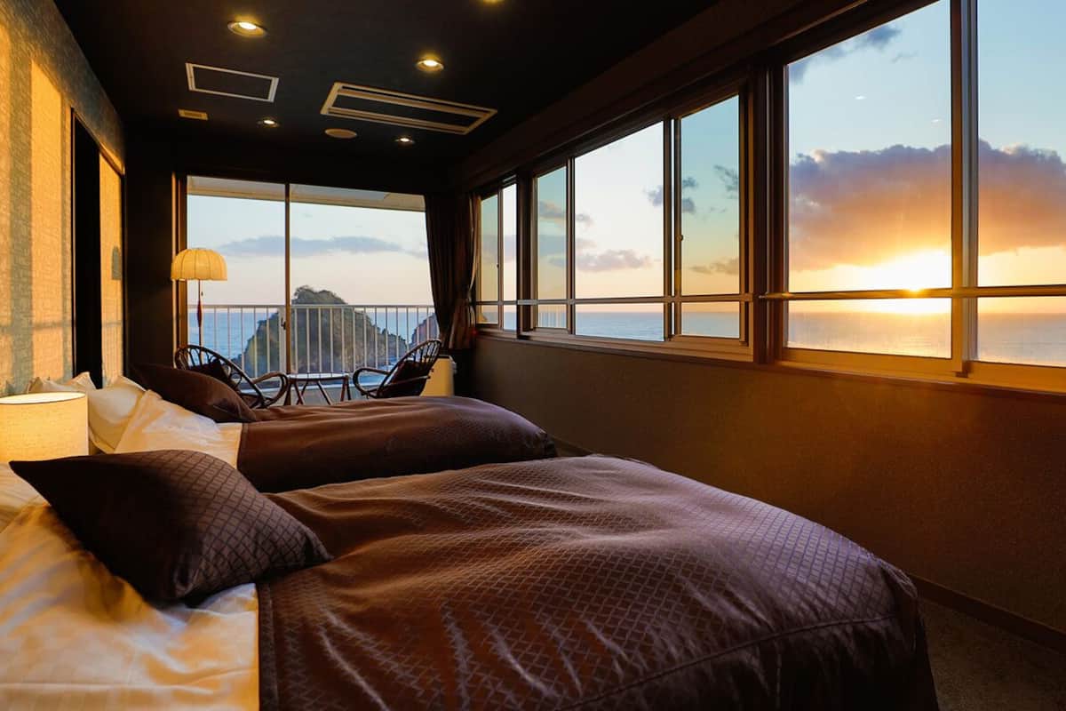 Quarto com cama, cadeira, abajur e uma grande janela com vista para o mar e para a cidade.
