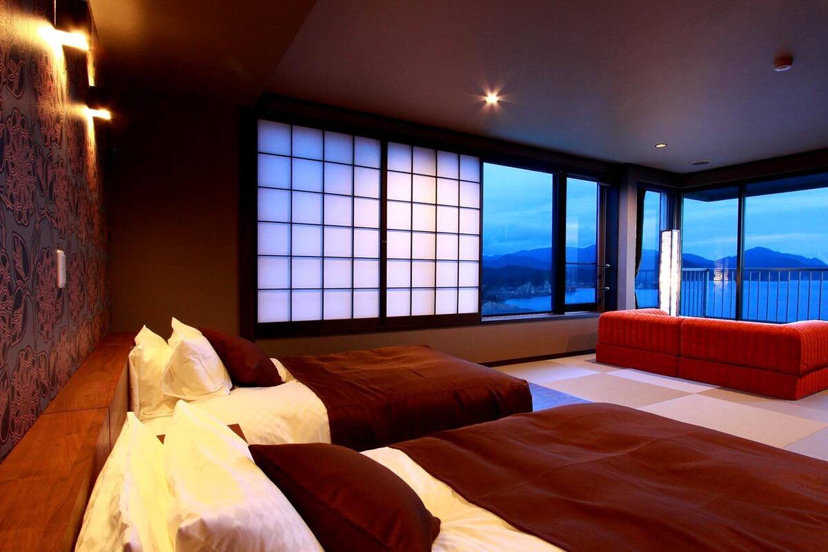 Quarto de hotel com duas camas, um sofá vermelho, janelas amplas com vista e parede estampada.