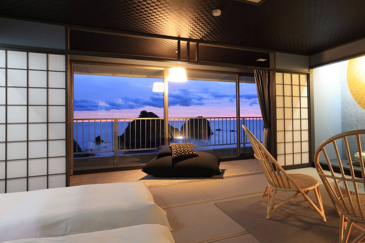 Quarto com cama, cadeira, mesa e vista para o mar ao entardecer.