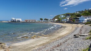 Uma praia com costa rochosa, um edifício branco com telhado vermelho e casas numa encosta.