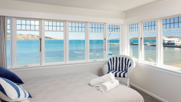 Quarto com grandes janelas dando para um corpo de água, uma cama com edredom branco, uma cadeira listrada e vista para um cais com barcos.