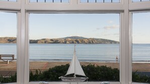Vista pela janela com um modelo de veleiro numa prateleira, um banco do lado de fora e uma praia com montanhas ao fundo.