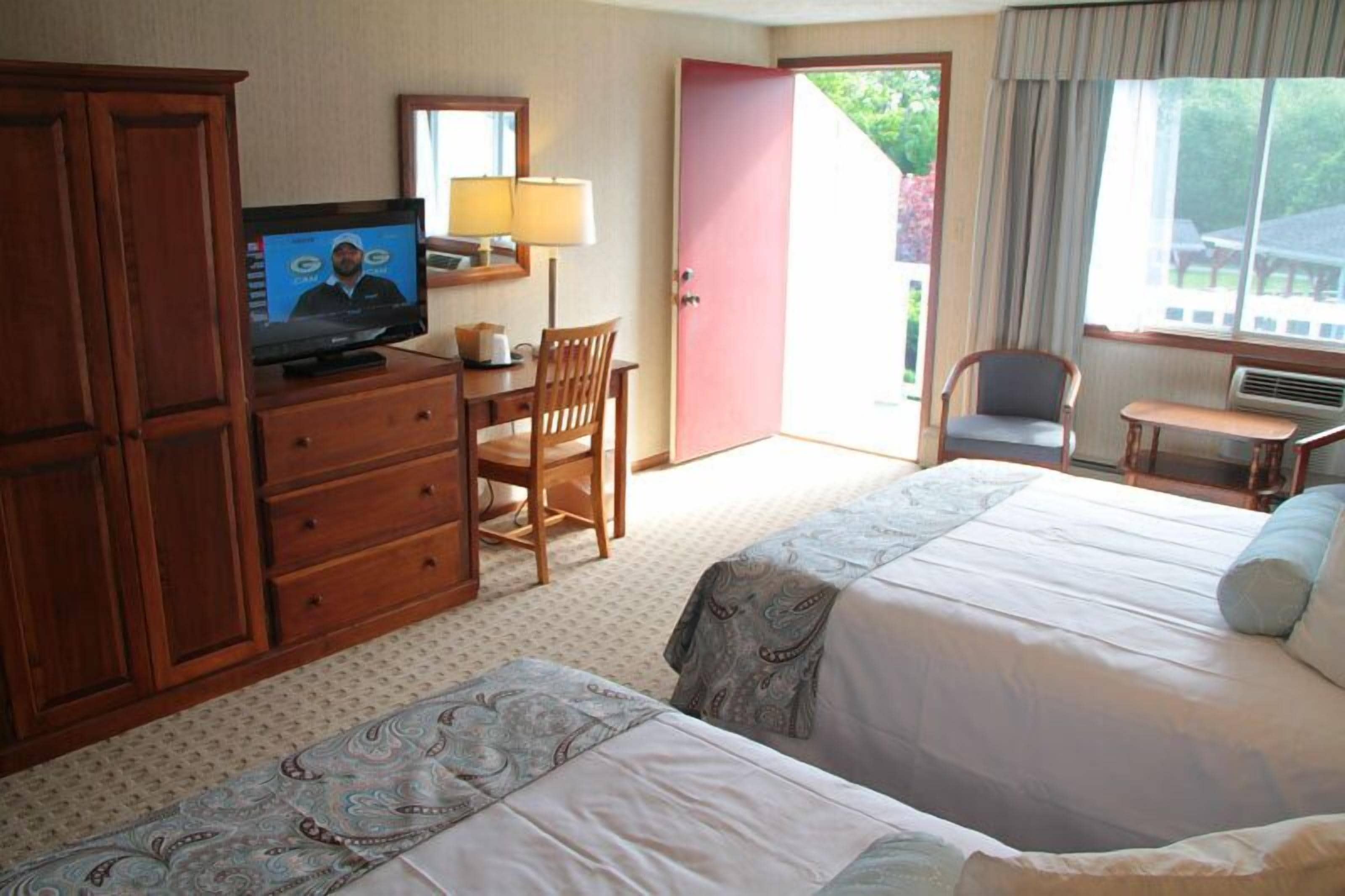Quarto de hotel com cama, escrivaninha com computador, cadeira, guarda-roupa e porta que dá para uma varanda.