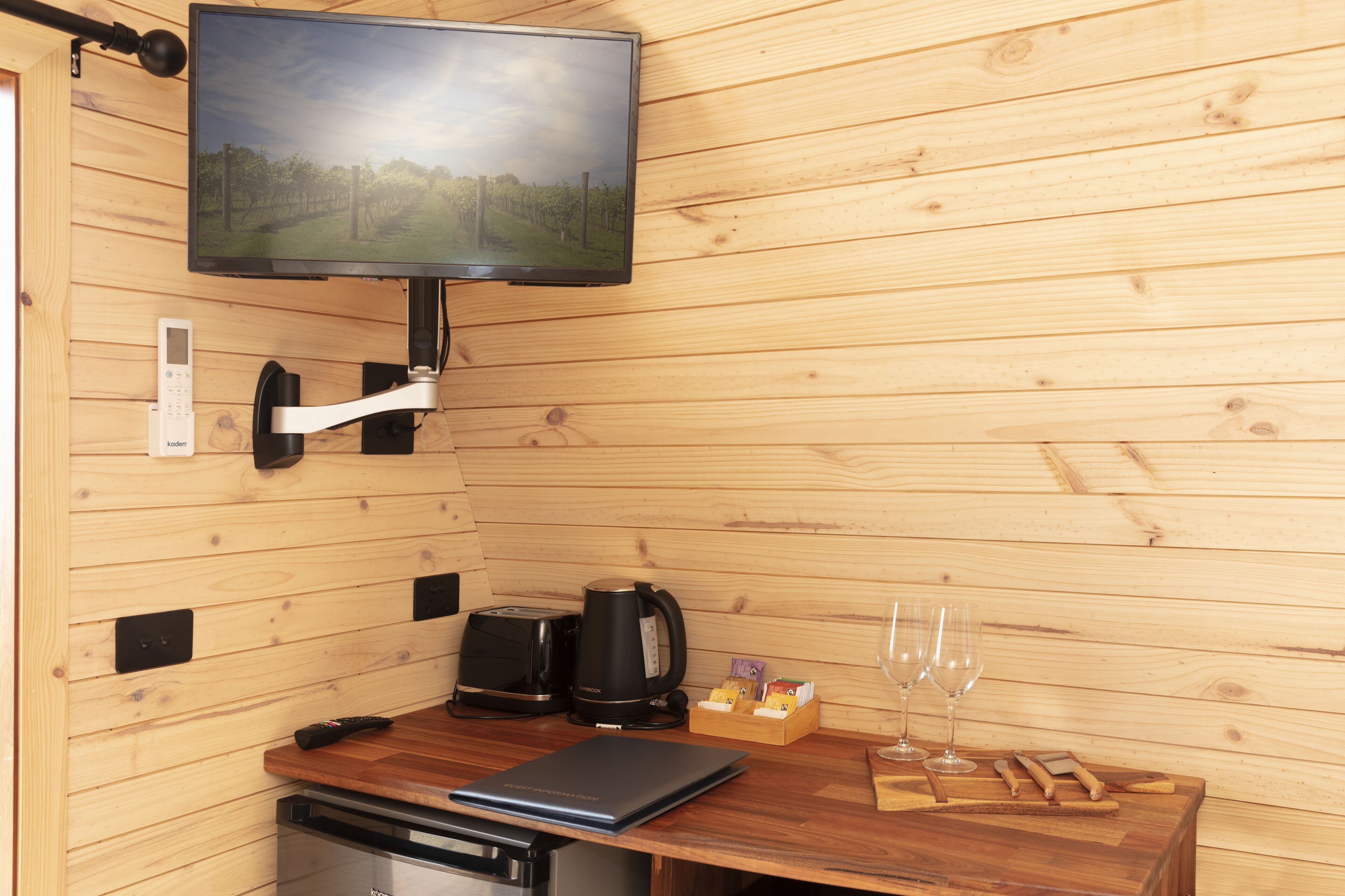 Interior de uma cabana de madeira com uma TV na parede, uma cafeteira, uma torradeira, um controle remoto, um laptop e um conjunto de copos de vinho.